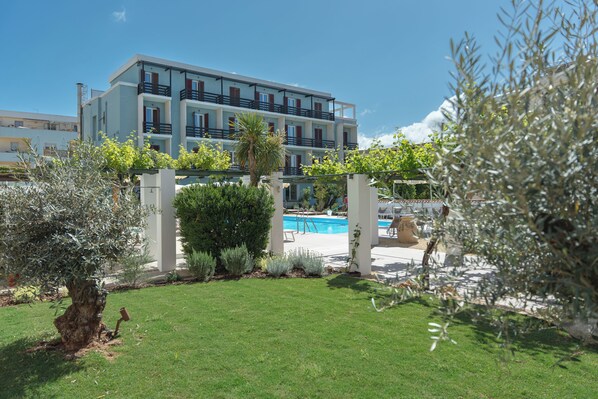Um edifício moderno com piscina, rodeado por vegetação bem cuidada e um céu claro.