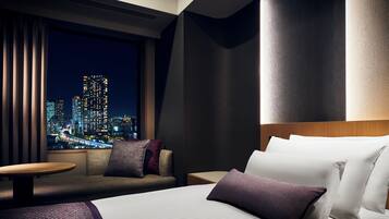 Quarto de hotel com cama, sofá, mesinha e vista noturna da cidade.