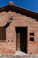 Um edifício rústico de tijolos com uma porta de madeira, uma pequena janela com portadas e um candeeiro na parede.