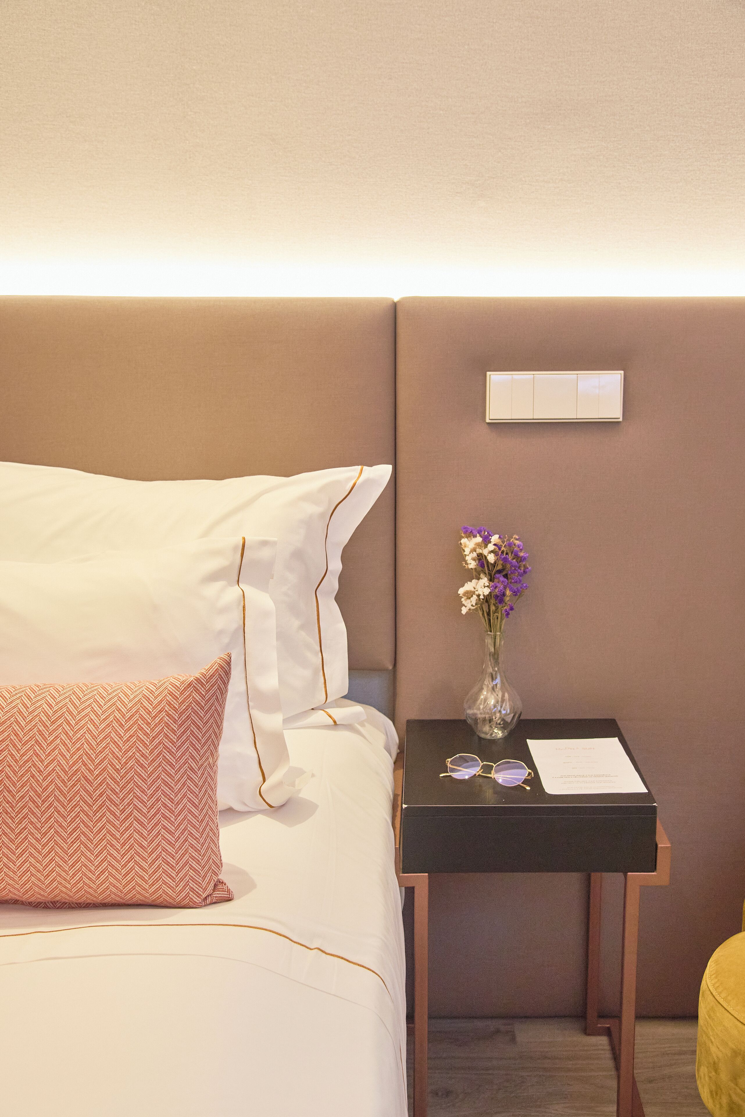 Cama bem arrumada com almofadas brancas e estampadas, uma pequena mesa de cabeceira com um vaso de flores e um interruptor de luz fixado na parede.
