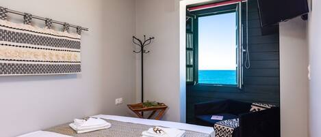 Quarto de hotel com uma cama, uma mesinha com uma planta, um cabideiro e vista para o mar através de uma porta aberta.