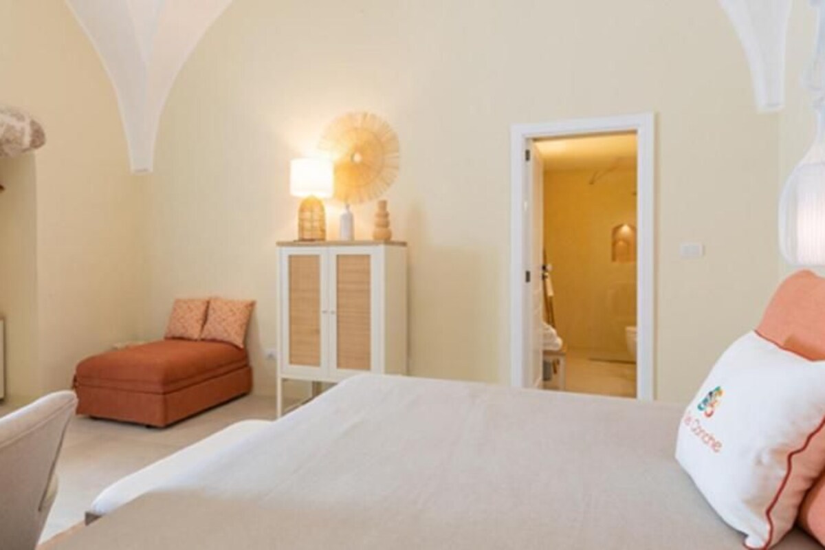 Um quarto com cama, uma cadeira, um armário e uma porta que dá para outro cômodo.