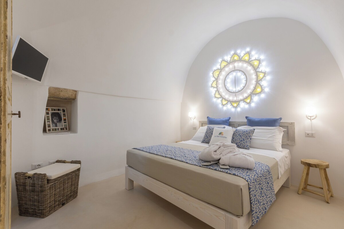 Quarto com cama, televisão na parede, luminária decorativa, um banquinho pequeno e uma cesta de vime.