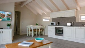Ampla sala de estar com mesa e cadeiras, cozinha com armários e eletrodomésticos brancos, e teto alto com vigas aparentes.