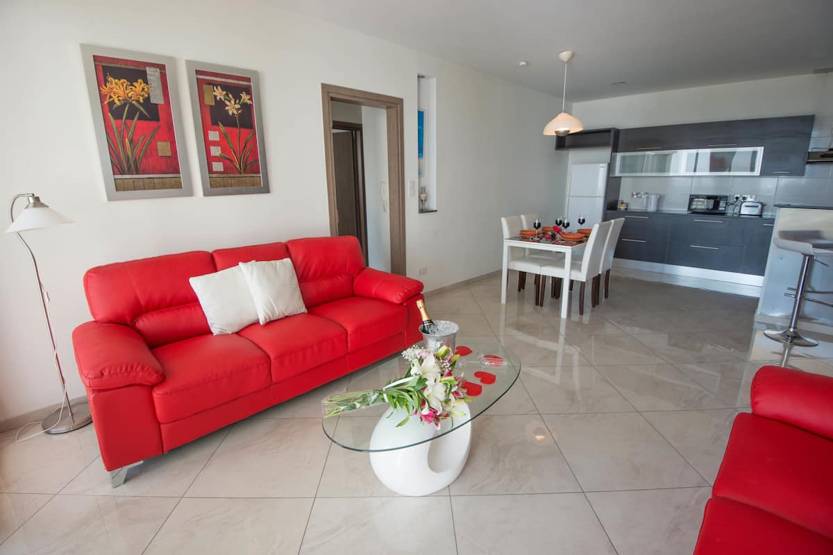 Uma sala de estar moderna com um sofá vermelho, uma mesa de centro de vidro com flores, uma área de jantar com mesa e cadeiras brancas, e uma cozinha com armários cinzentos.