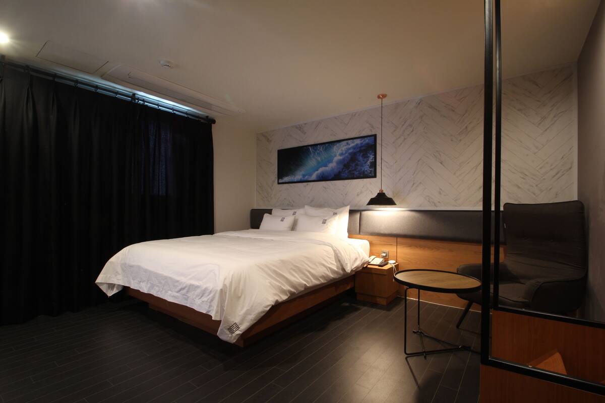 Quarto de hotel moderno com uma cama grande, uma mesa de cabeceira, abajur, cadeira e uma janela ampla.