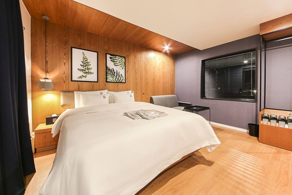 Quarto moderno com teto de madeira, uma cama grande, mesa de cabeceira, uma cadeira, janela com persianas e um sapateiro.