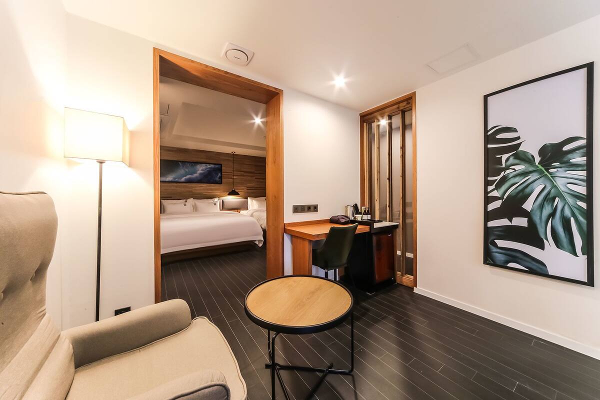 Quarto de hotel com cama, escrivaninha, cadeira, luminária e uma planta grande na parede.