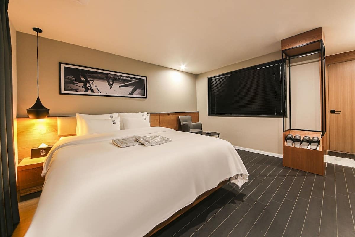 Quarto de hotel com uma cama grande, uma mesa de cabeceira com luminária, uma cadeira, uma janela com persianas e um guarda-roupa de madeira.