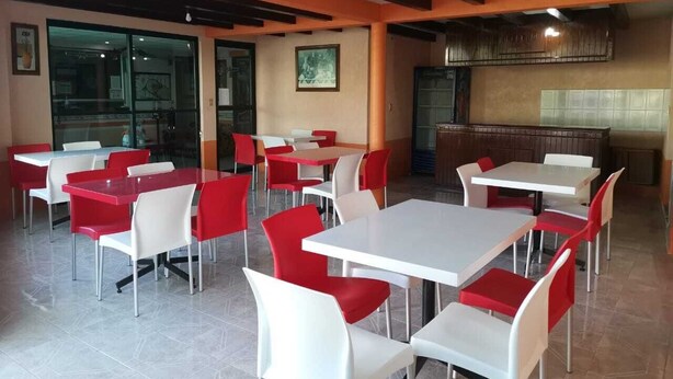 Um restaurante com mesas e cadeiras vermelhas e brancas, uma área de cozinha e janelas amplas.