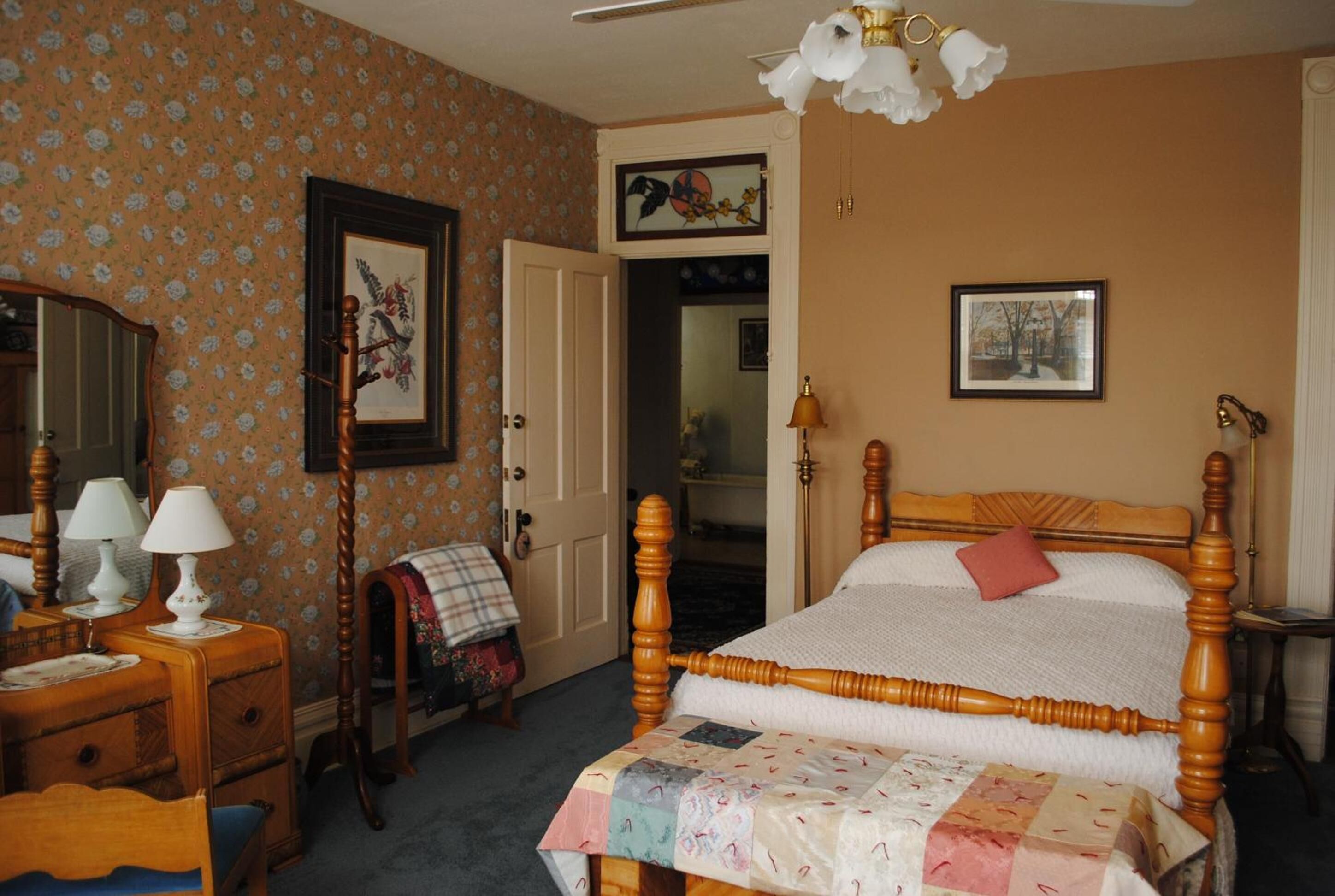 Um quarto com uma cama de madeira baldaquino, uma mesa de cabeceira com luminárias, um aparador e uma porta que dá para outro cômodo.