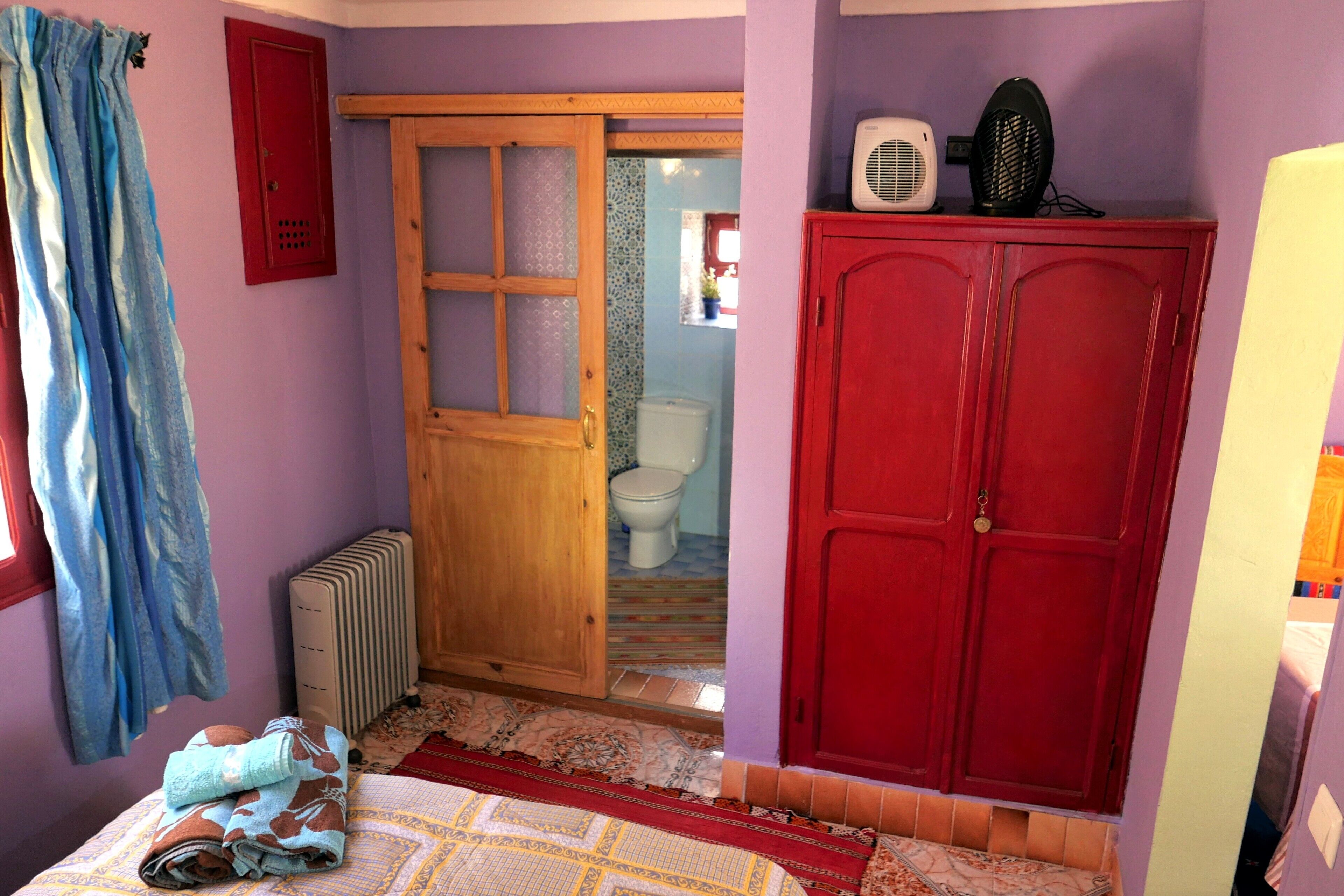 Quarto com cama, guarda-roupa, radiador, janela com cortinas e porta de acesso a um banheiro.