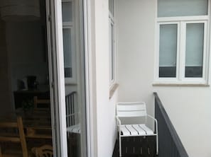 Uma pequena varanda com uma cadeira branca e uma mesa, uma janela e uma porta que dá para um quarto com uma planta e uma cafeteira.
