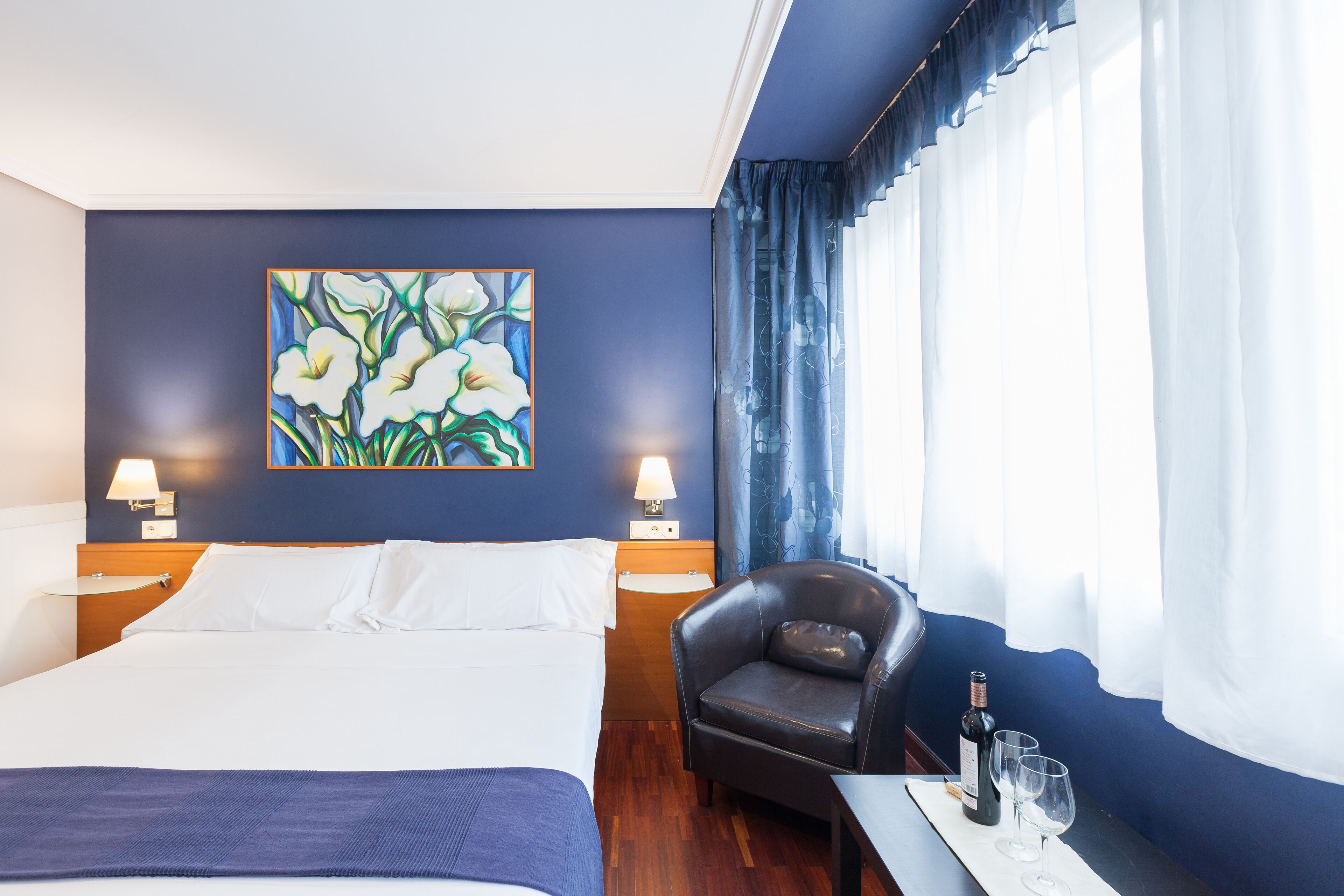 Quarto de hotel com uma cama grande, uma cadeira, uma mesa com garrafa e copos, e um quadro na parede.