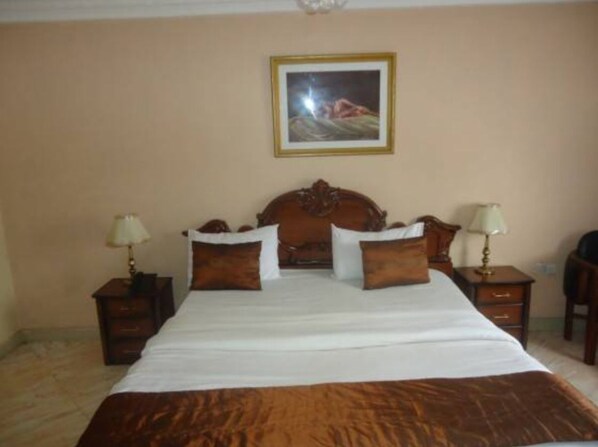 Cama bem arrumada com cabeceira de madeira, duas mesas de cabeceira com abajures e um quadro pendurado na parede.