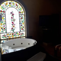 Um banheiro com uma banheira branca, uma grande janela de vitrais com motivos florais e uma televisão na parede.