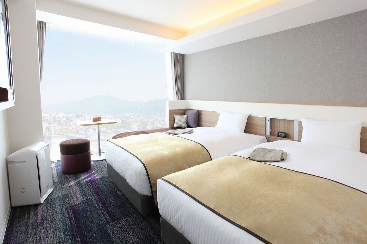 Quarto de hotel com duas camas, uma mesinha, uma cadeira e vista para a cidade.