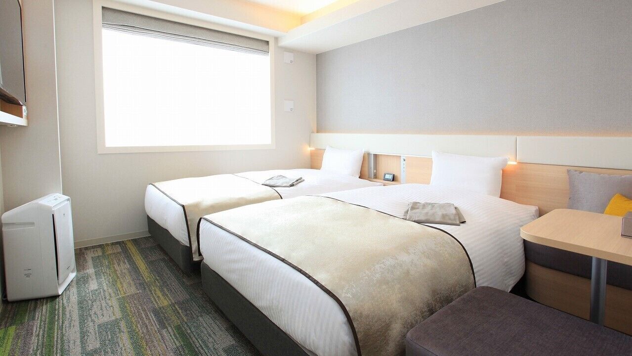 Quarto de hotel com duas camas, uma escrivaninha, uma cadeira, uma mesinha, um aquecedor e uma janela.