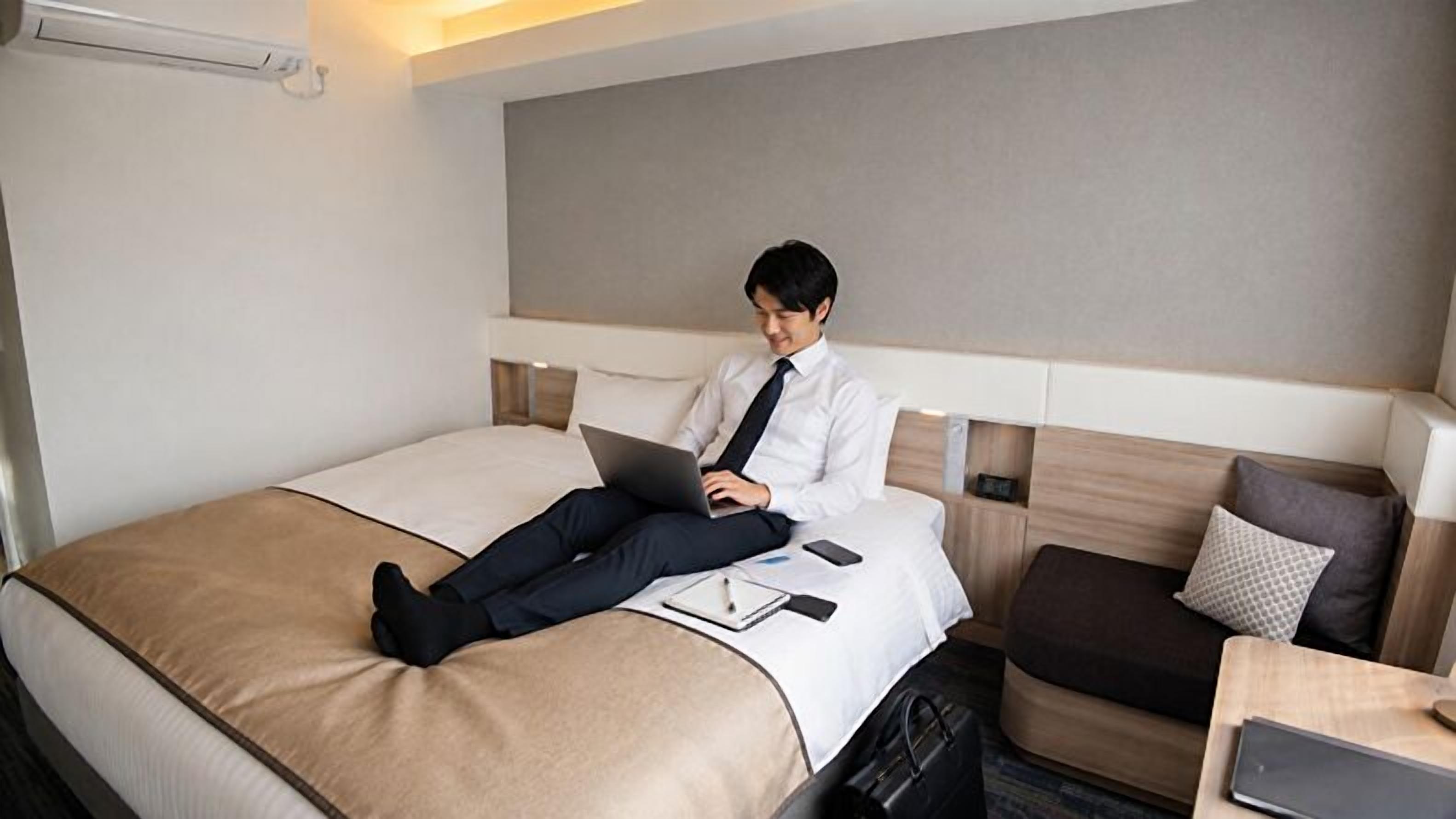 Quarto de hotel com cama, escrivaninha, laptop e uma pessoa trabalhando.