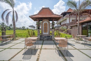 Um pavilhão tradicional com telhado de azulejos, rodeado por área externa com assentos de vime e mesas, com um fundo de campo verdejante e céu límpido.
