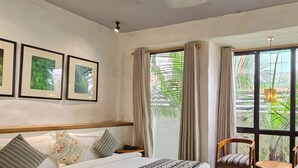 Quarto de hotel com cama, duas cadeiras, escrivaninha com luminária e janelas amplas com cortinas.