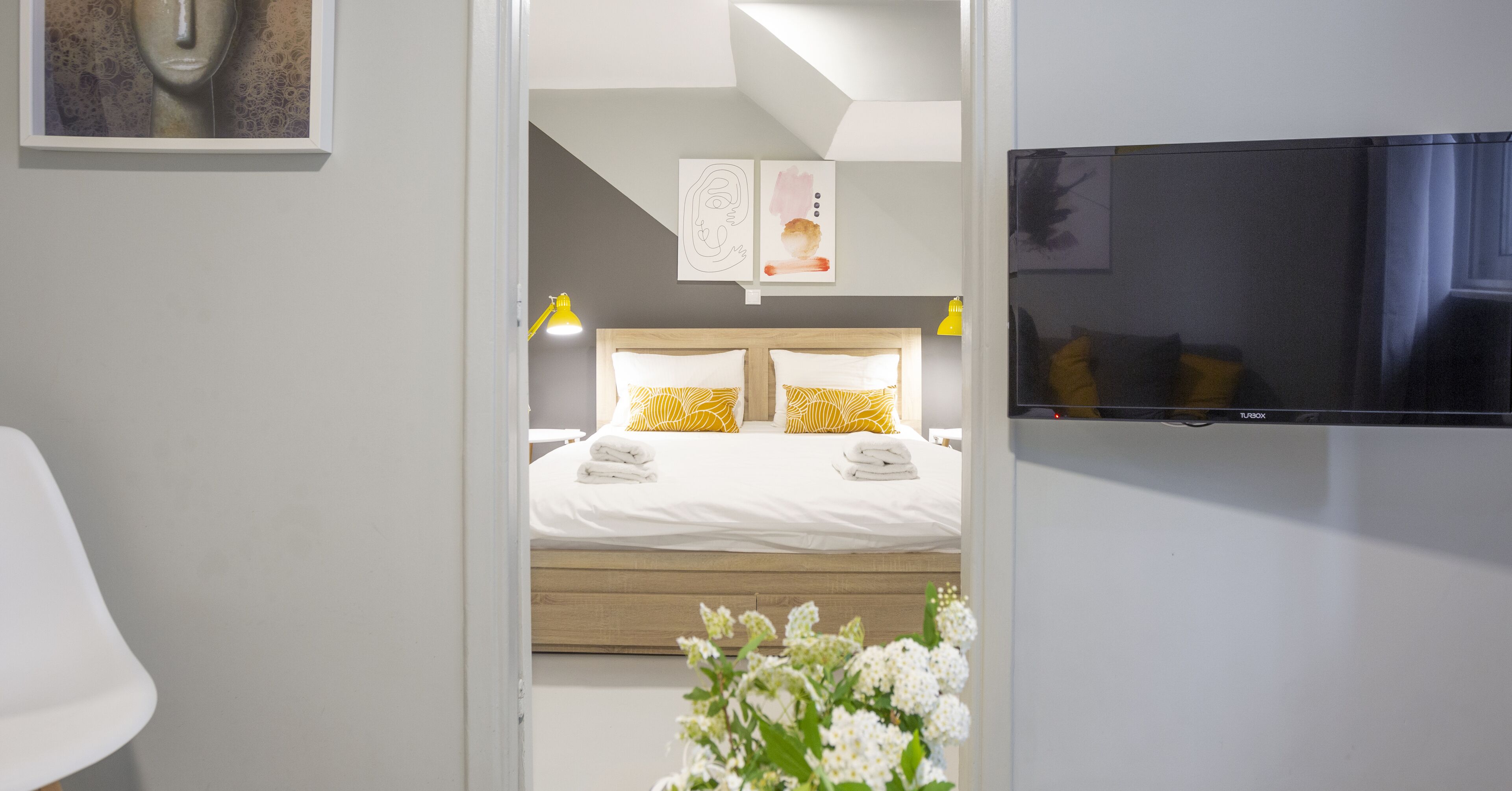 Quarto de hotel com cama, duas luminárias laterais, televisão de tela plana e uma obra de arte emoldurada na parede.