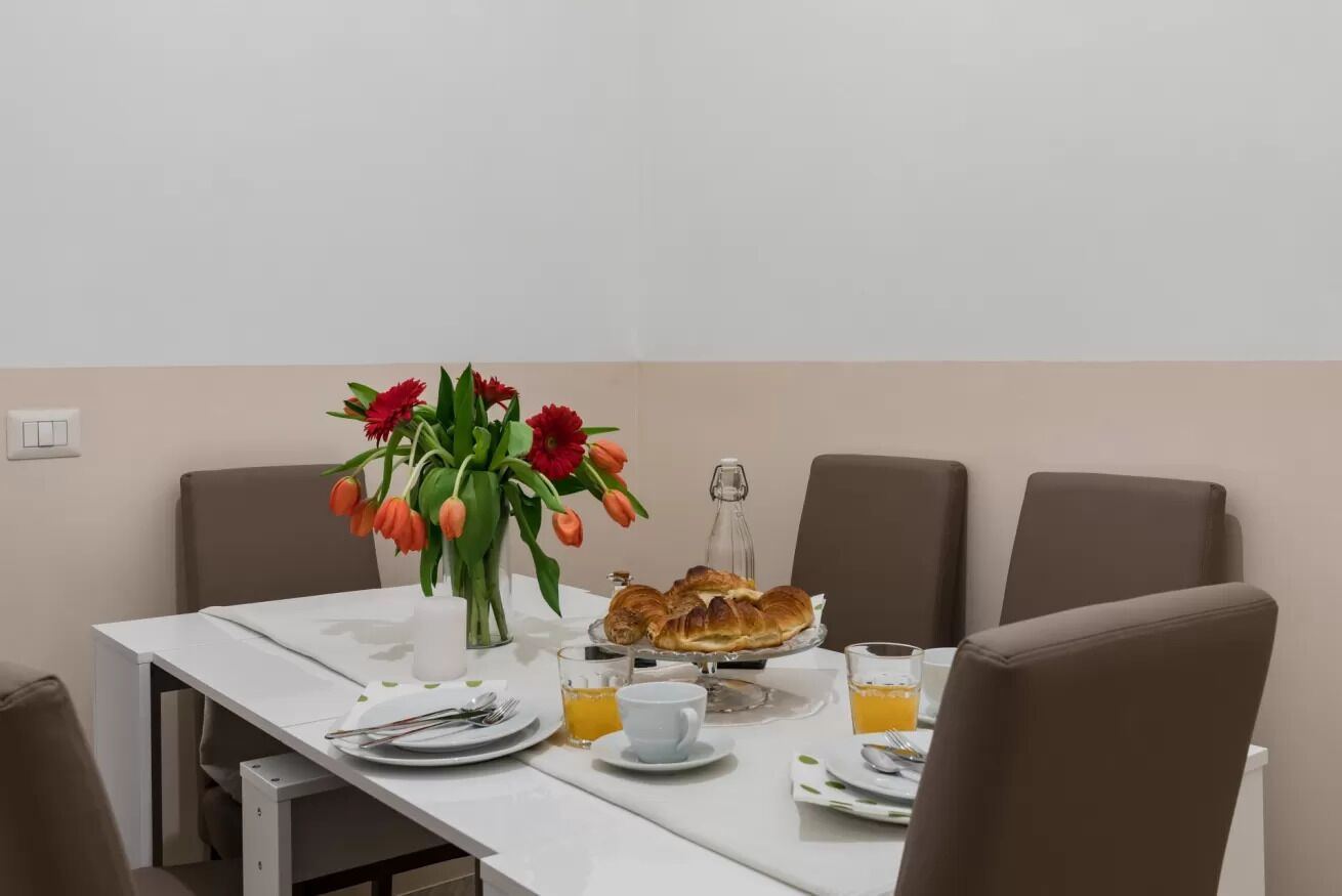 Uma mesa de jantar posta com pratos, talheres, copos, um vaso de flores e um croissant.