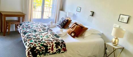 Quarto de hotel com cama, escrivaninha, cadeira, luminária e janela com cortinas.