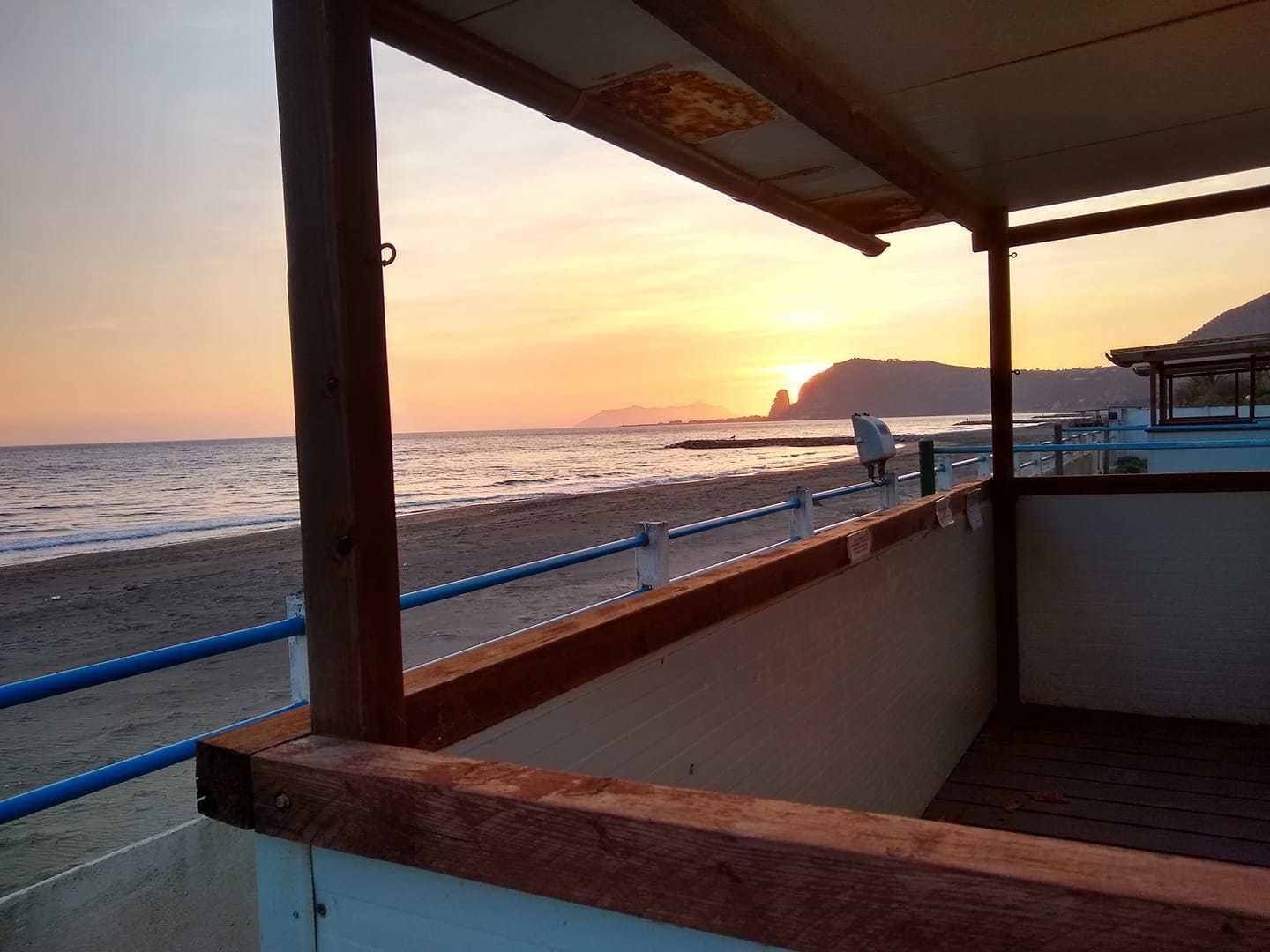 Vista da praia a partir de uma área coberta com guarda-corpo, um deck de madeira e um pôr do sol sobre o oceano.