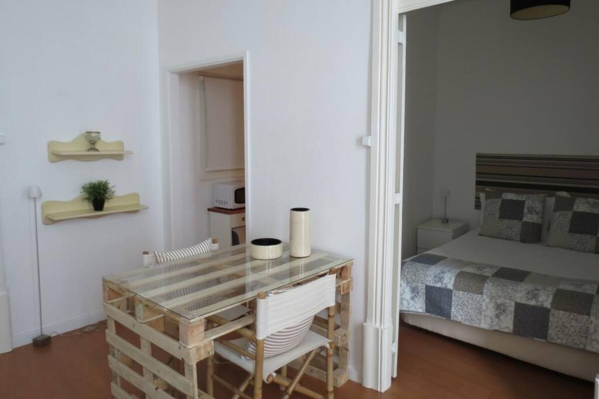 Um quarto pequeno e bem iluminado, com uma mesa e cadeira de madeira, uma cama com colcha xadrez e uma estante com uma planta e uma vela.