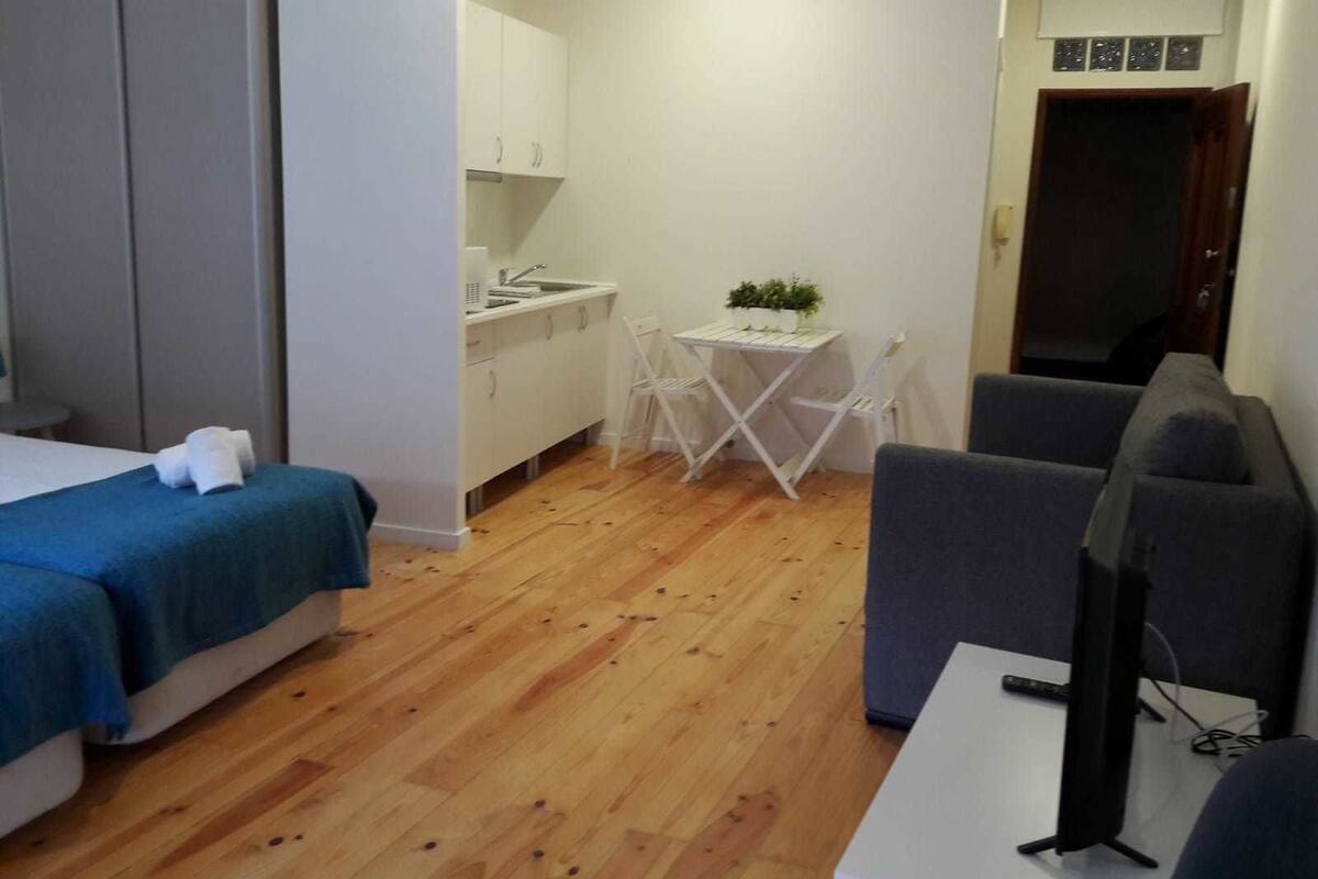 Quarto compacto com uma cama, pequeno espaço de cozinha, mesa de jantar com cadeiras e televisão.