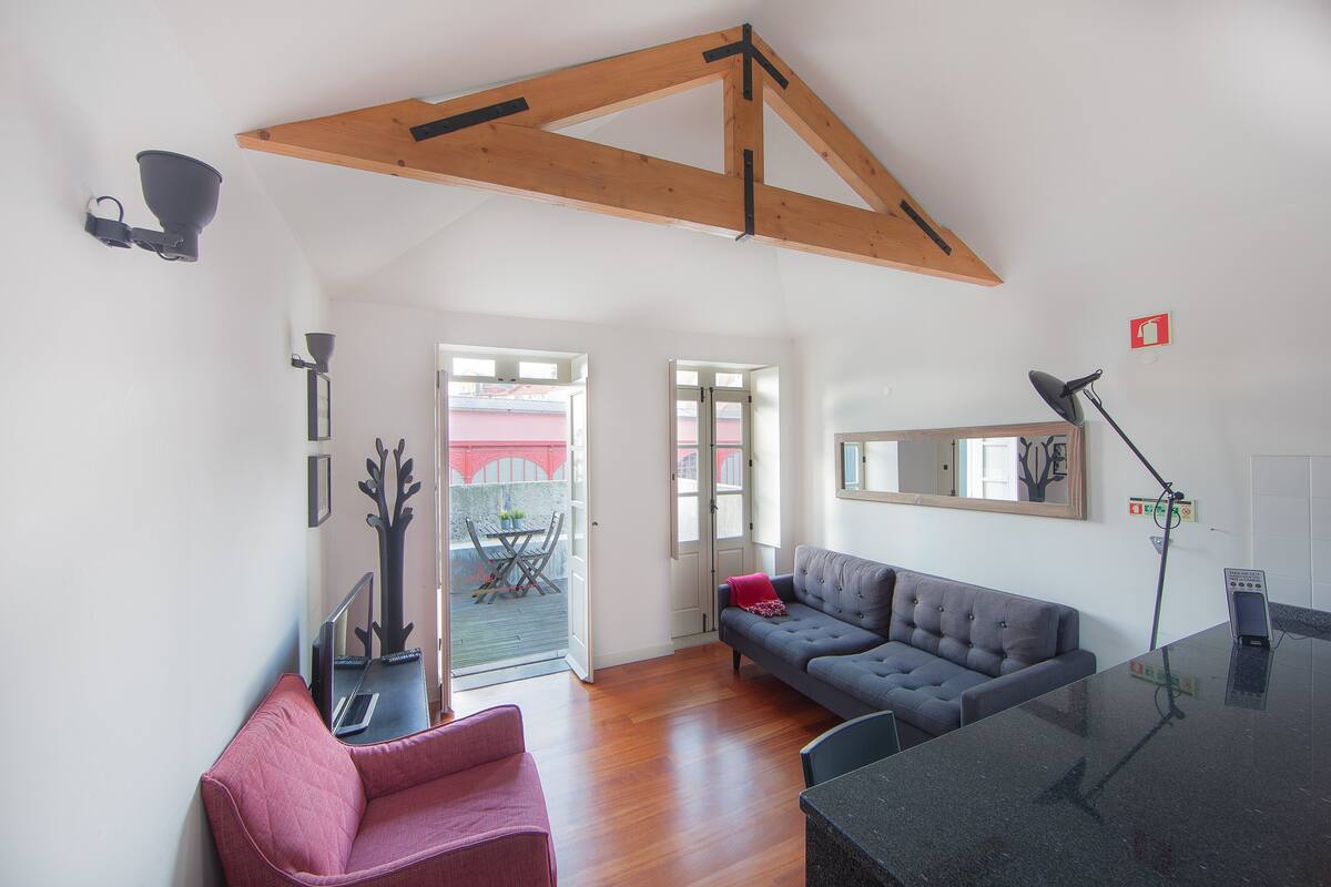 Uma sala de estar ampla com teto de madeira, um sofá cinzento, uma poltrona rosa e uma mesa de vidro.