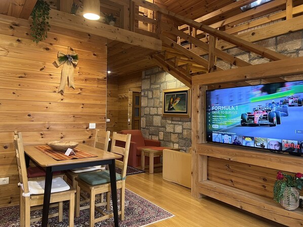 Interior acolhedor de uma cabana de madeira, com área de jantar, televisão exibindo uma corrida de Fórmula 1 e uma parede de pedra.