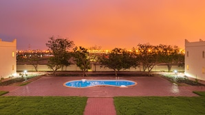 Uma piscina rodeada por um caminho de tijolos, com árvores e vista para edifícios distantes ao pôr do sol.