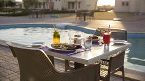 Mesa de jantar com pratos, talheres e bebidas à beira da piscina.