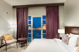 Quarto de hotel com cama, uma cadeira, uma mesinha e vista para a praia à noite.