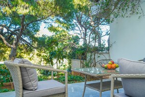 Uma área de estar externa com móveis de vime e mesa de vidro, cercada por vegetação e árvores.