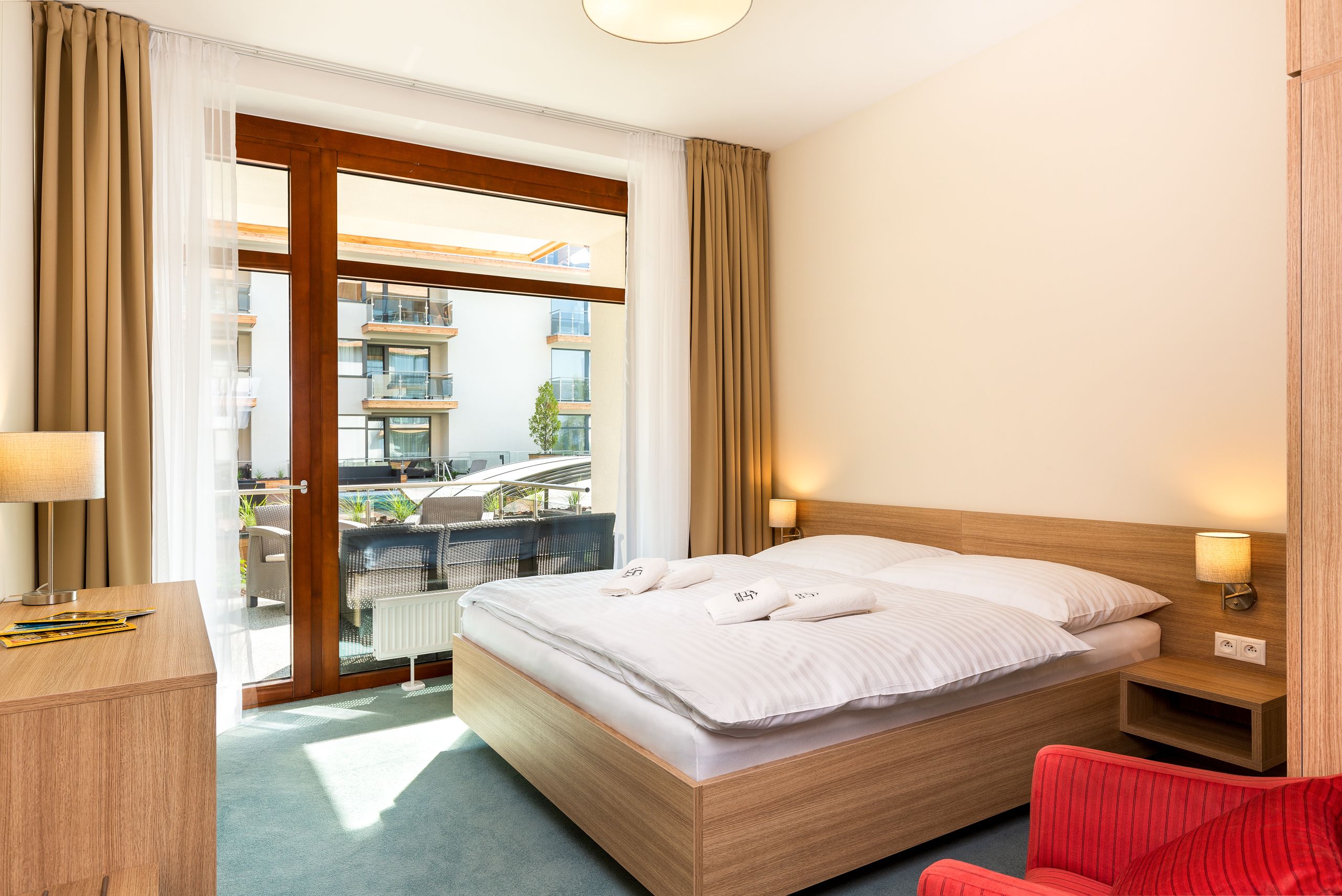 Quarto de hotel com cama de madeira, uma escrivaninha, uma cadeira, um abajur e vista para a varanda.