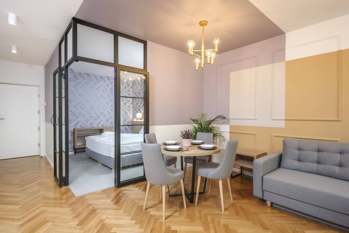 Quarto moderno com divisória de vidro, uma cama, mesa de jantar com cadeiras, sofá e lustre.