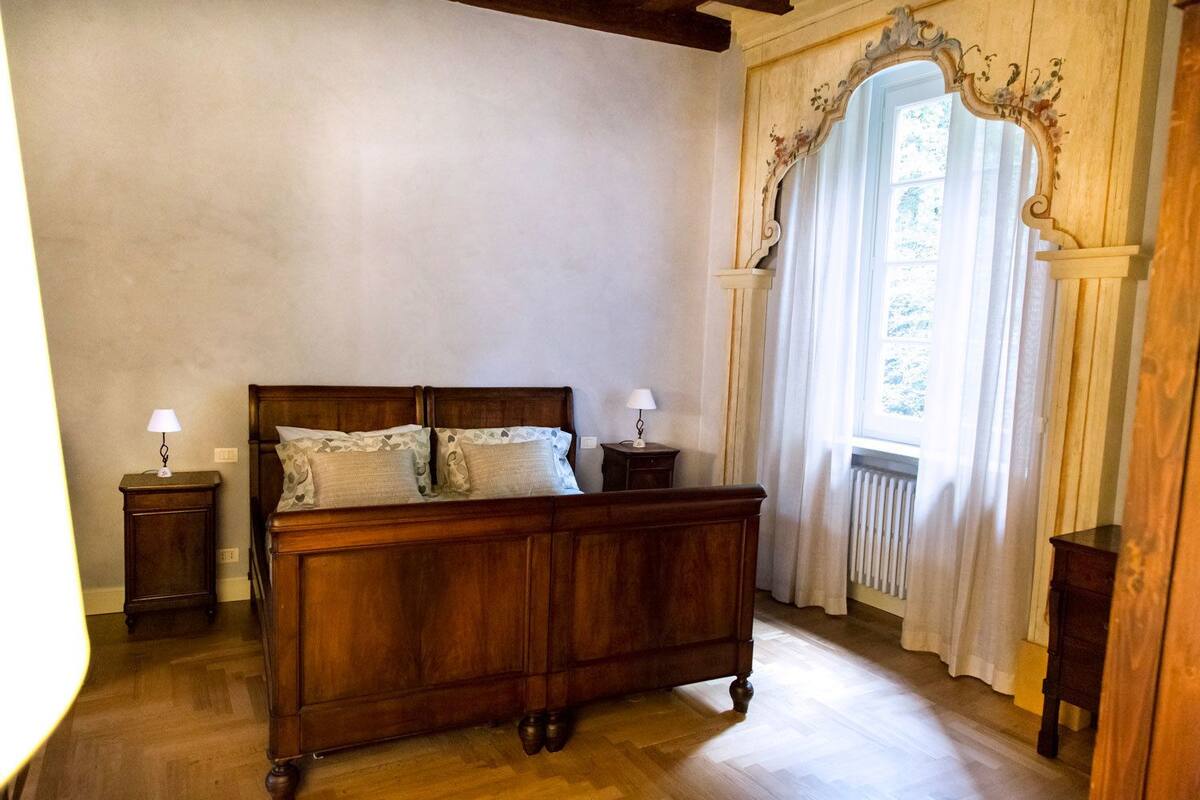 Quarto com cama de madeira, duas mesas de cabeceira, uma janela com cortinas e um radiador.