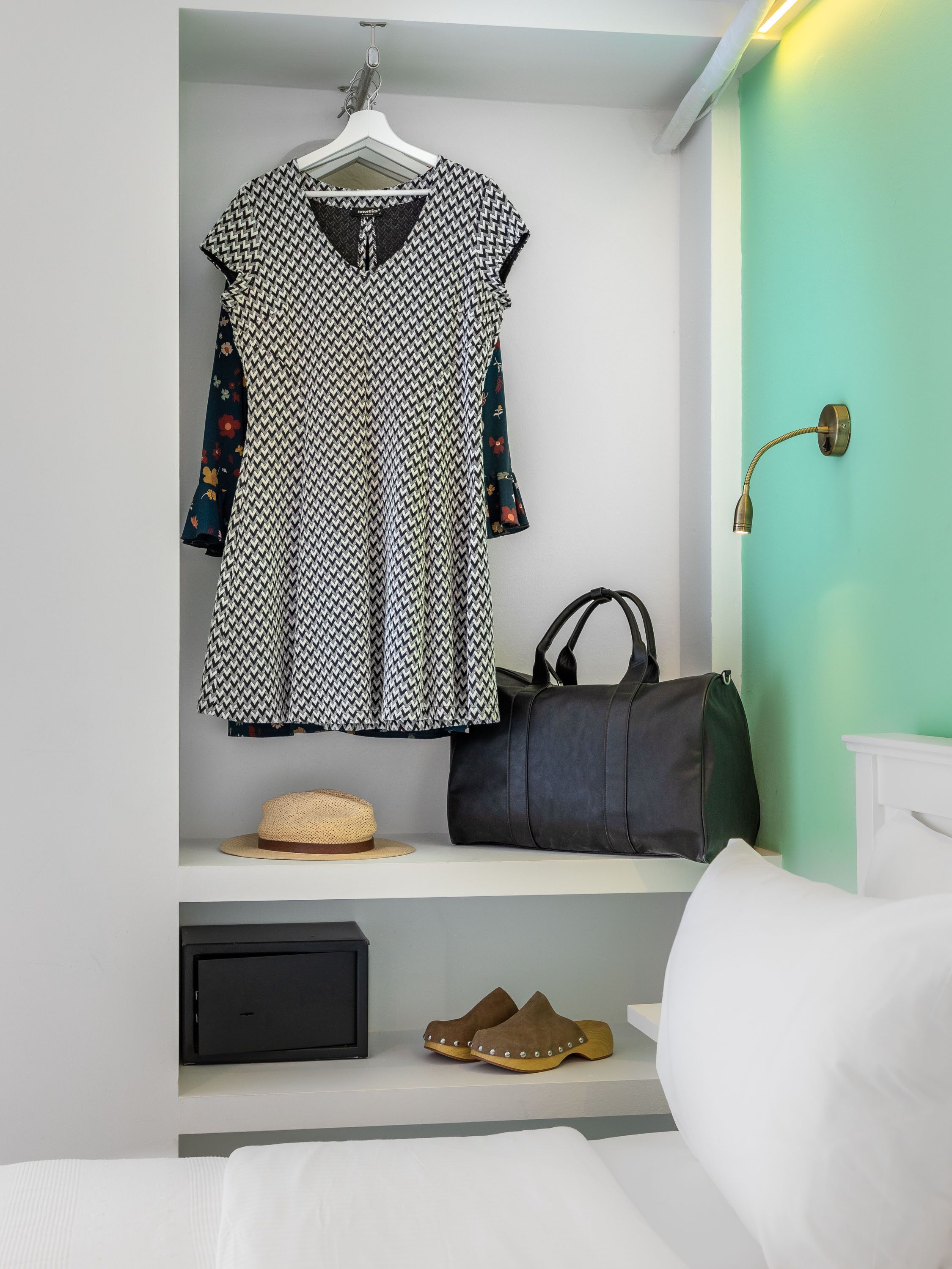 Quarto bem organizado com um vestido pendurado, uma bolsa, um chapéu, uma mala e um par de sapatos numa estante.