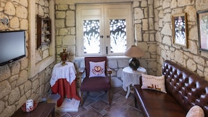 Um quarto com paredes de pedra, teto de madeira, um candelabro, televisão, sofá, poltrona, uma mesinha e um tapete estampado.