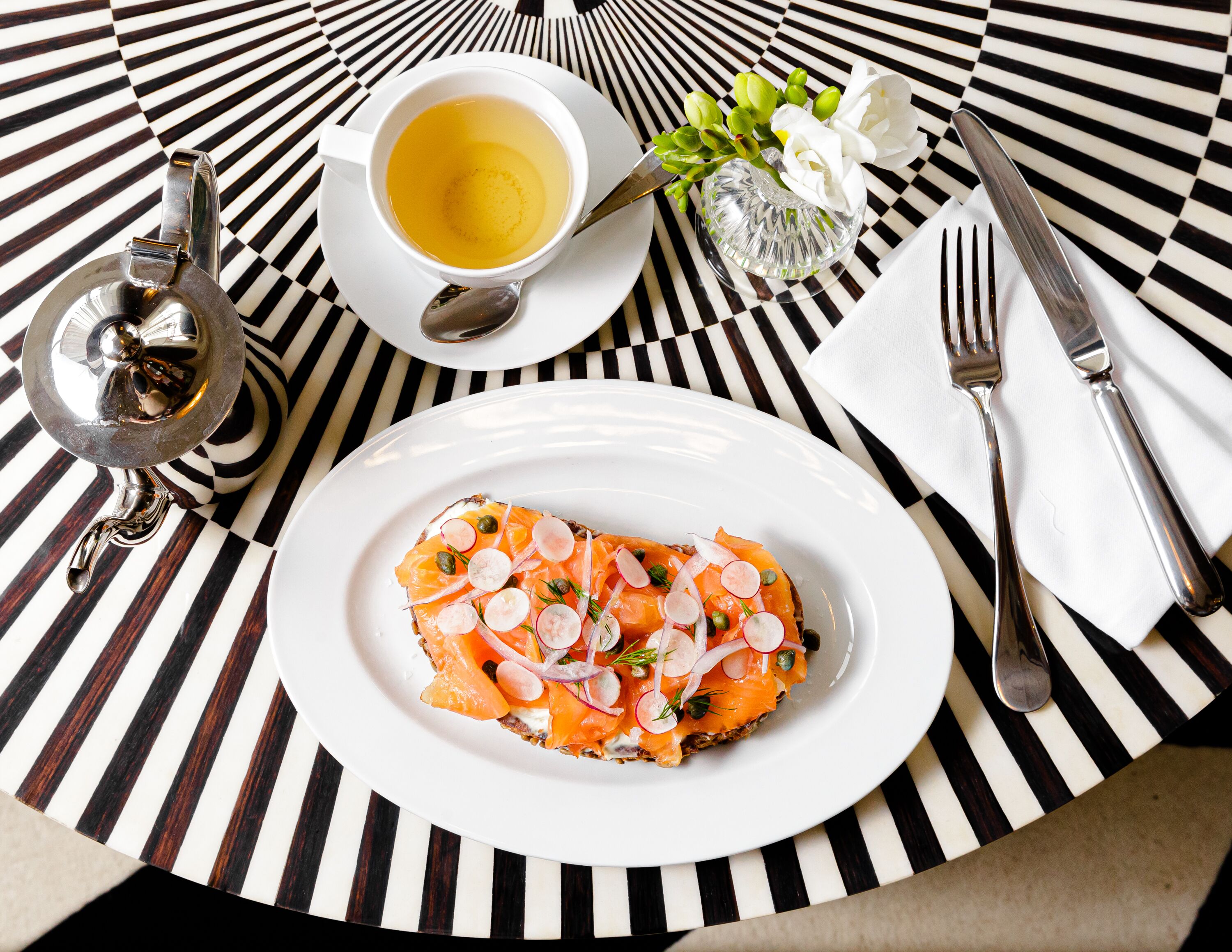 Uma mesa com toalha listrada, um prato branco com salmão e rabanetes, um vaso de vidro transparente com flores brancas, uma chaleira e talheres sobre um guardanapo.