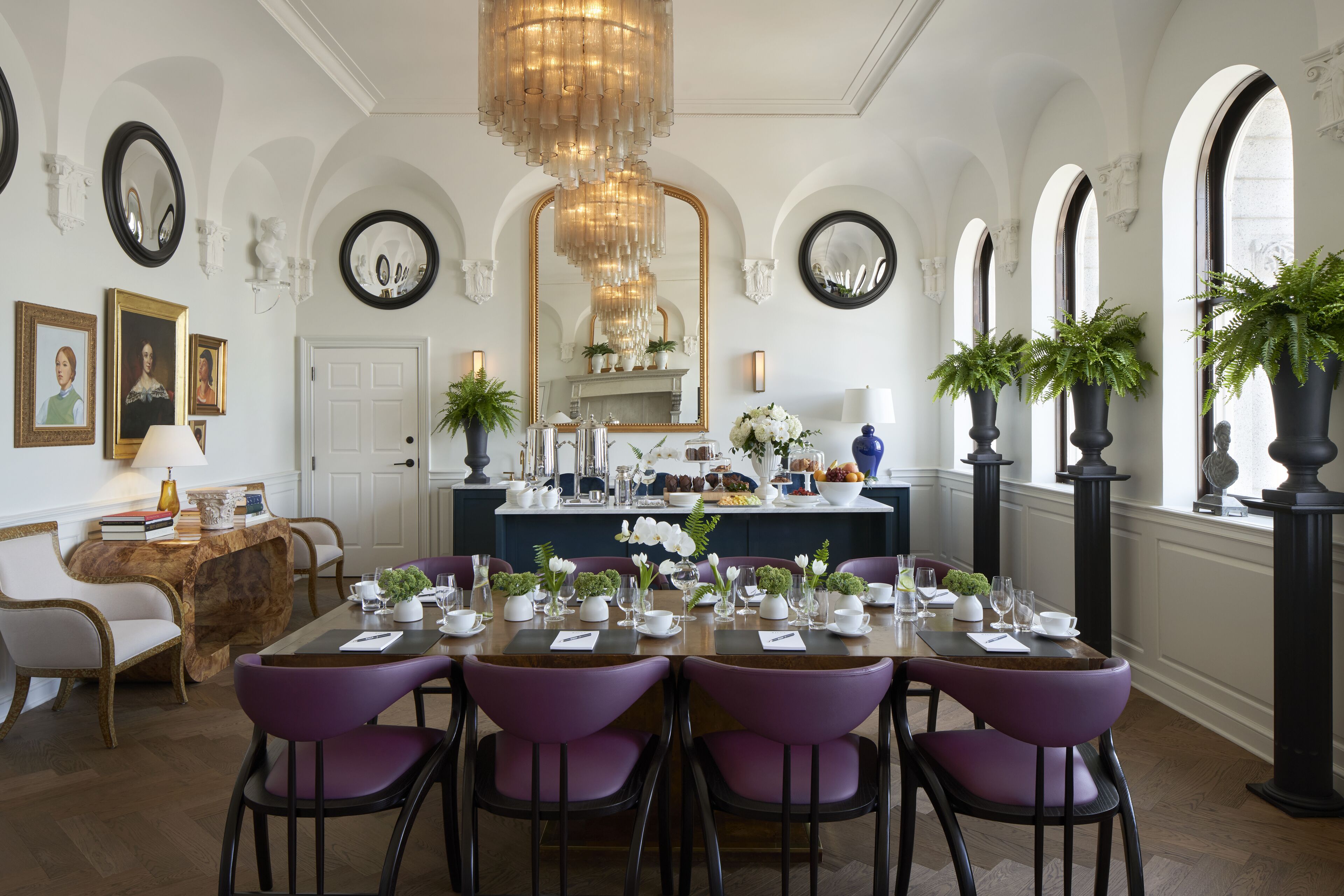 Uma sala de jantar com uma mesa comprida posta para seis pessoas, cercada por cadeiras roxas. Há plantas em vasos no parapeito da janela e um lustre sobre a mesa.