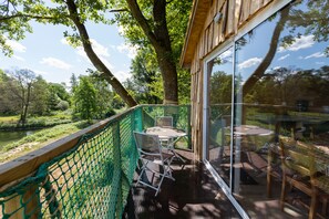 Um alpendre externo envidraçado com mesa e cadeiras, cercado por uma cerca verde, com vista para um rio e árvores.