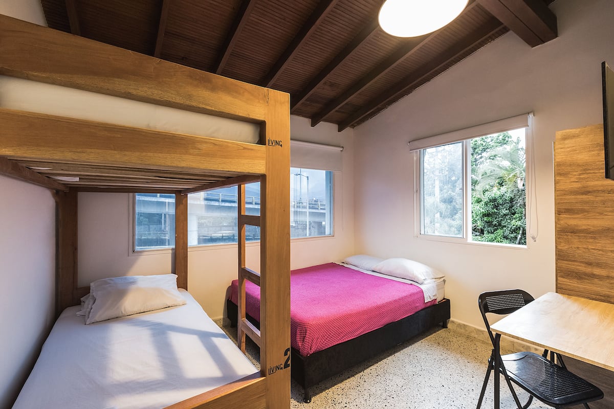 Quarto com cama de solteiro, cama individual, escrivaninha e cadeira. Janelas oferecem luz natural e vista para o exterior.