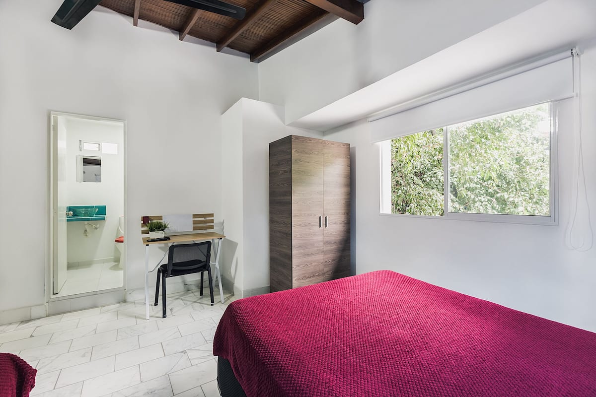 Quarto com cama, escrivaninha e guarda-roupa. Uma janela permite entrada de luz natural.