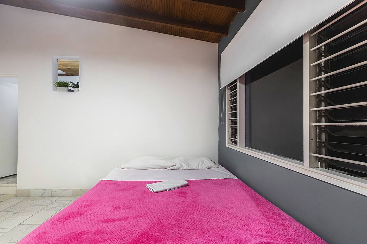 Quarto com cama, janela com persianas, uma pequena planta na parede e porta que dá para outro cômodo.