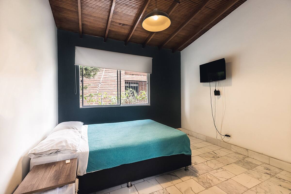 Quarto com cama, janela, televisão fixada na parede e teto de madeira.