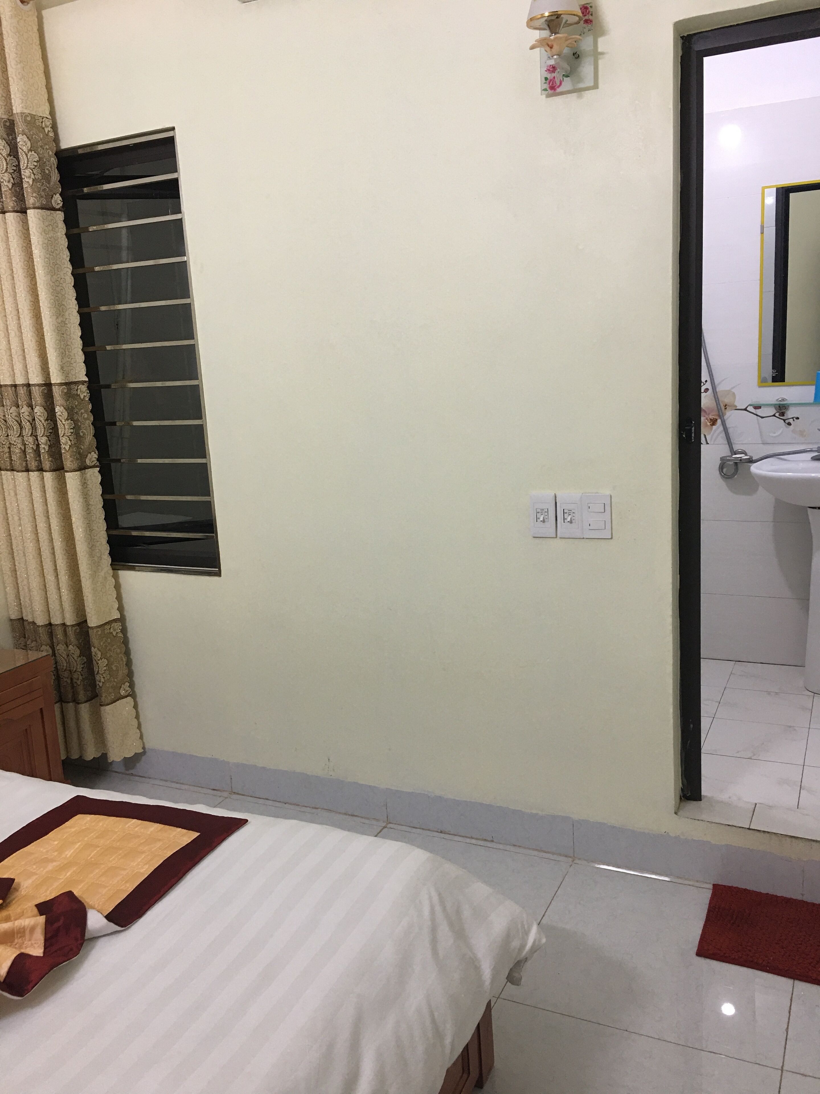 Quarto com cama, janela com cortinas, luminária fixada na parede e banheiro visível através de uma porta aberta.